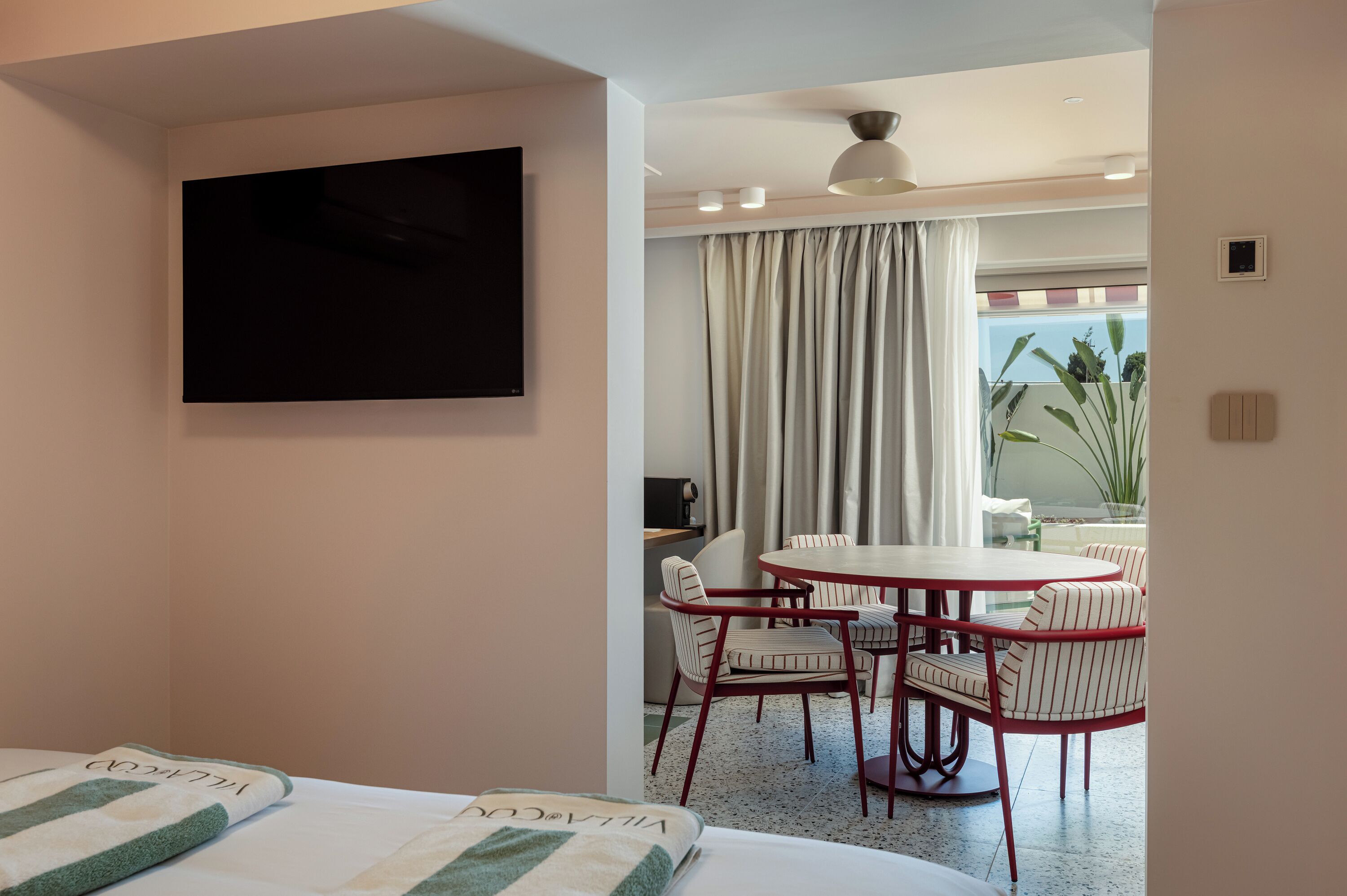 Quarto de hotel com cama, televisão presa na parede, área de refeições com mesa e cadeiras, e vista para o exterior.