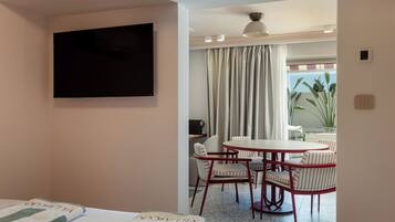 Quarto de hotel com cama, televisão presa na parede, área de refeições com mesa e cadeiras, e vista para o exterior.