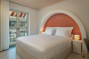 Quarto de hotel com uma cama grande, uma mesa de cabeceira com abajur e vista para uma varanda com mobiliário.