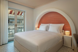 Quarto de hotel com uma cama grande, uma mesa de cabeceira com abajur e vista para uma varanda com mobiliário.