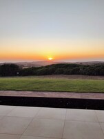 Vista do pôr do sol a partir de um terraço pavimentado, com um campo gramado e colinas ao longe.
