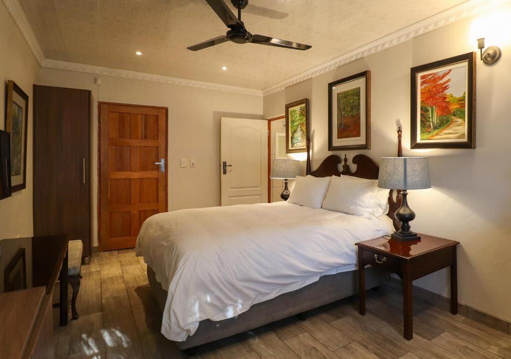 Quarto de hotel com cama, mesa de cabeceira, abajur, ventilador de teto e obras de arte nas paredes.