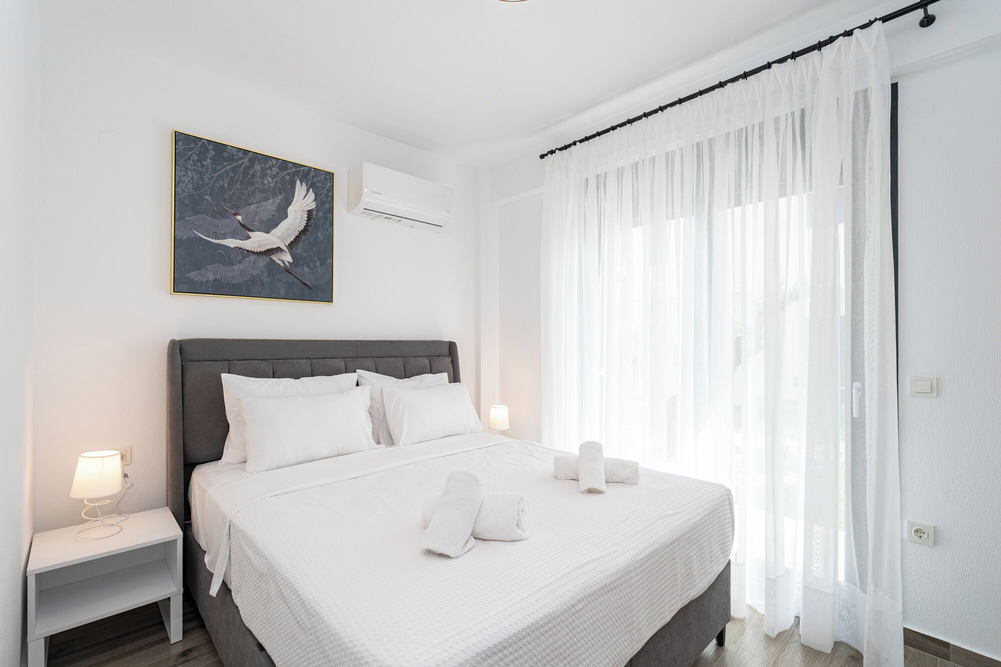 Quarto de hotel com cama, roupa de cama branca, mesa de cabeceira com abajur, pintura de pássaros e cortinas finas.