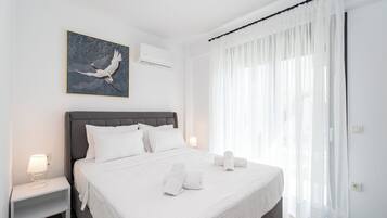 Quarto de hotel com cama, roupa de cama branca, mesa de cabeceira com abajur, pintura de pássaros e cortinas finas.