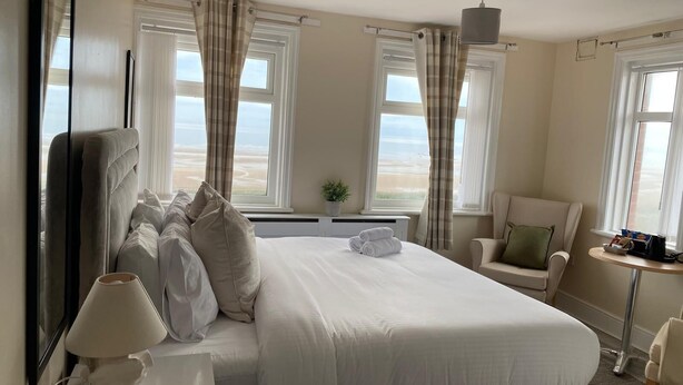 Quarto de hotel com uma cama grande, uma cadeira, uma mesinha e vista para a praia.