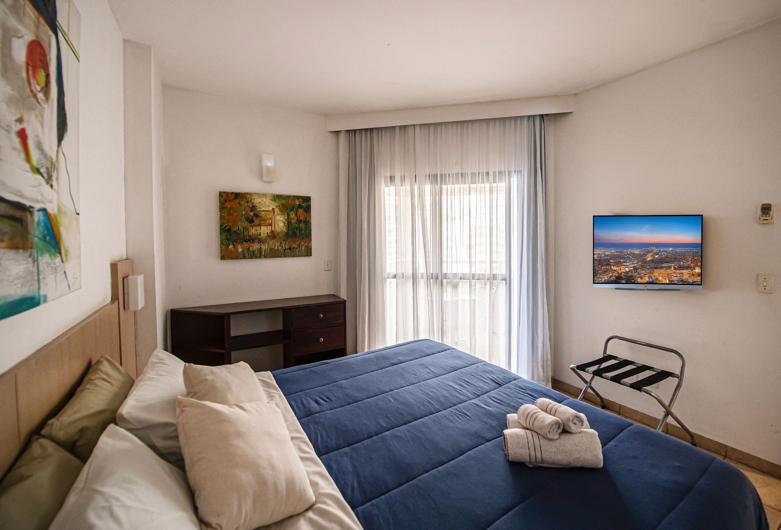 Quarto de hotel com uma cama, uma mesa, uma cadeira, um quadro e uma televisão.