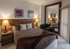 Quarto de hotel com uma cama grande, duas mesas de cabeceira com abajures, um sofá pequeno e um espelho amplo.
