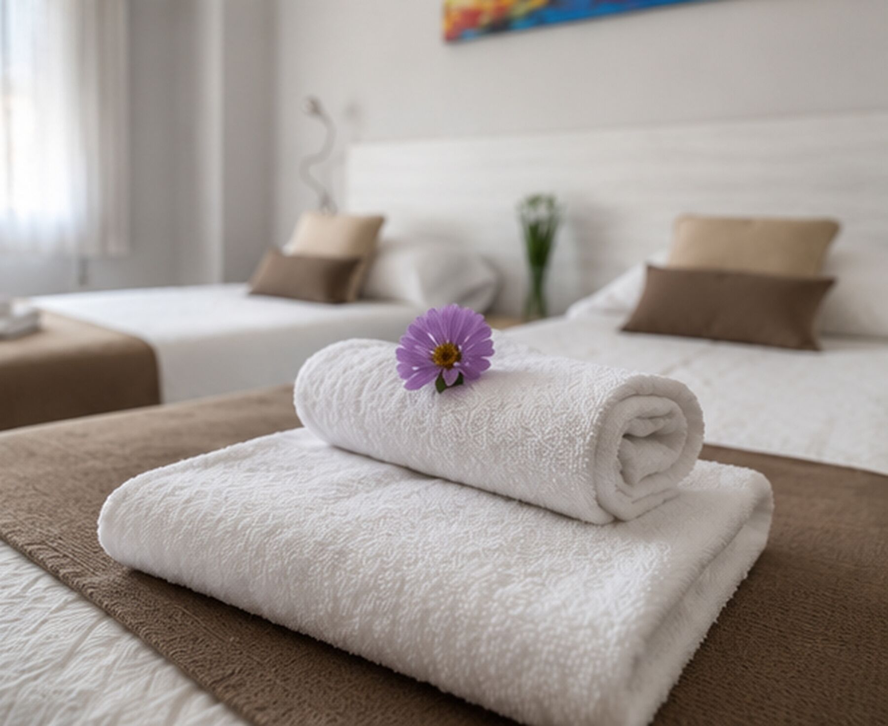 Quarto de hotel com duas camas, cada uma com uma almofada castanha e cabeceira branca. Uma toalha branca enrolada está colocada sobre uma toalha castanha na cama.