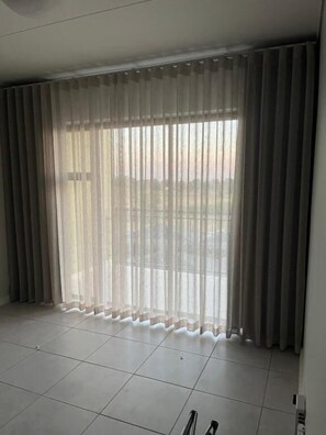 Quarto com uma grande janela coberta por cortinas leves, piso de azulejos e maçaneta visível.