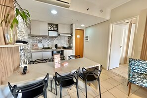Cozinha moderna com armários brancos, painel de azulejos e porta de madeira. Há uma mesa de jantar com quatro cadeiras e uma pequena planta à esquerda.
