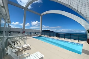 Uma área de piscina com espreguiçadeiras, céu claro e vista para o oceano.