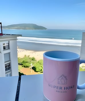 Uma caneca rosa com a inscrição 'SUPER HOST' e 'BOSTON', colocada numa varanda com vista para uma praia, ondas e uma colina ao longe.