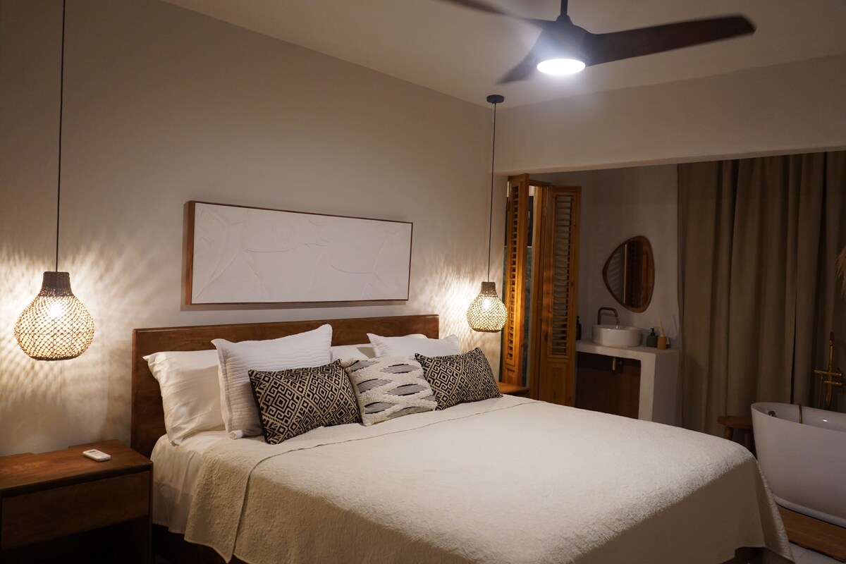 Uma cama bem arrumada com almofadas estampadas, uma mesa de cabeceira com abajur e um ventilador de teto.