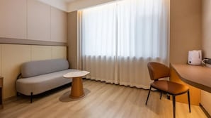 Quarto de hotel moderno com sofá, uma mesa redonda pequena, uma cadeira, uma janela grande com cortinas e uma área de kitchenette.