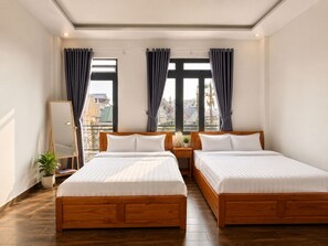 Quarto de hotel com duas camas de madeira, um espelho grande, um vaso de planta e vista para edifícios através de amplas janelas.