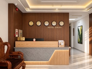 Bancada de recepção de hotel com parede de painel de madeira, vários relógios, placa de recepção e área de espera com uma cadeira.