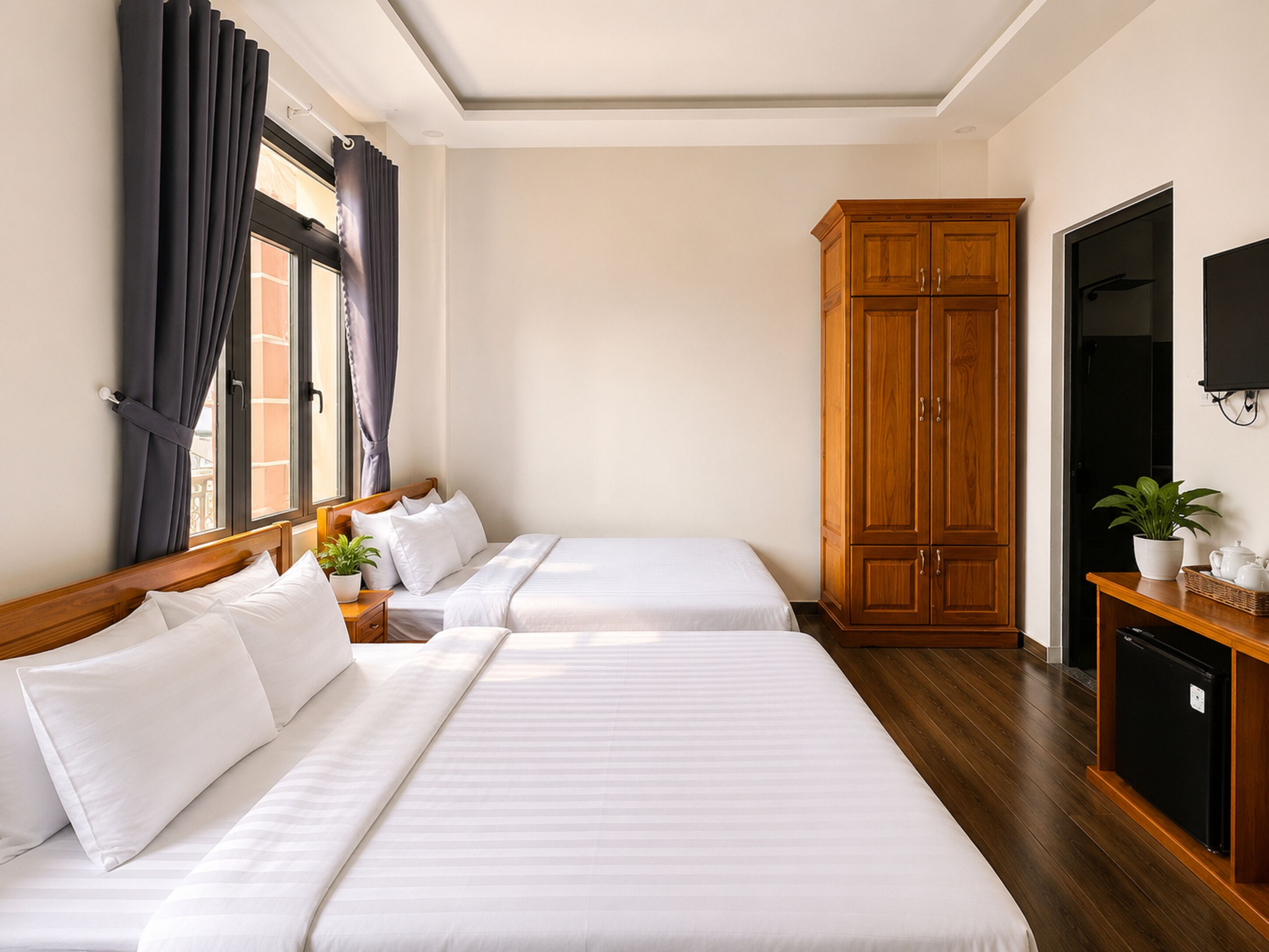 Quarto de hotel com duas camas, um guarda-roupa, uma televisão e uma pequena mesa com uma planta.