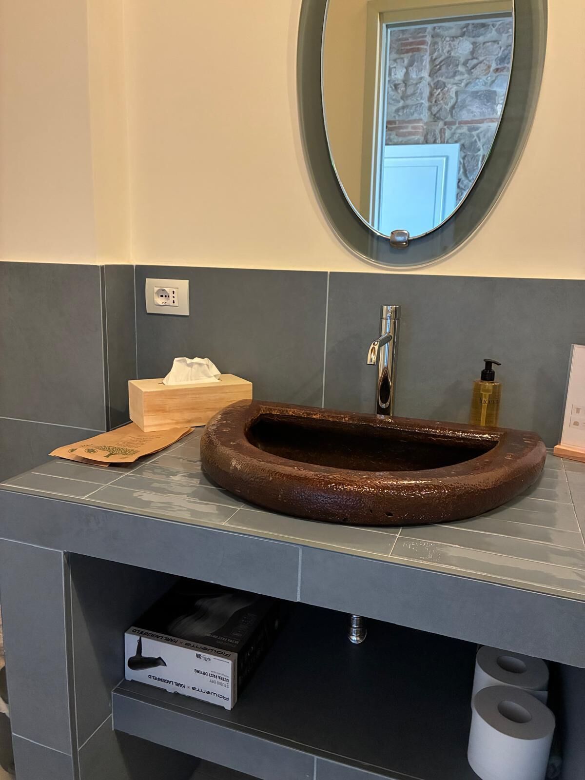 Uma casa de banho com uma pia redonda e escura, um espelho, uma caixa de lenços e um frasco de sabonete líquido.