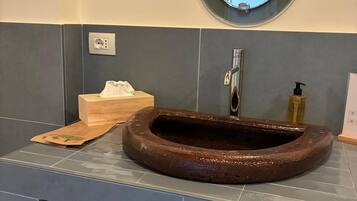 Uma casa de banho com uma pia redonda e escura, um espelho, uma caixa de lenços e um frasco de sabonete líquido.