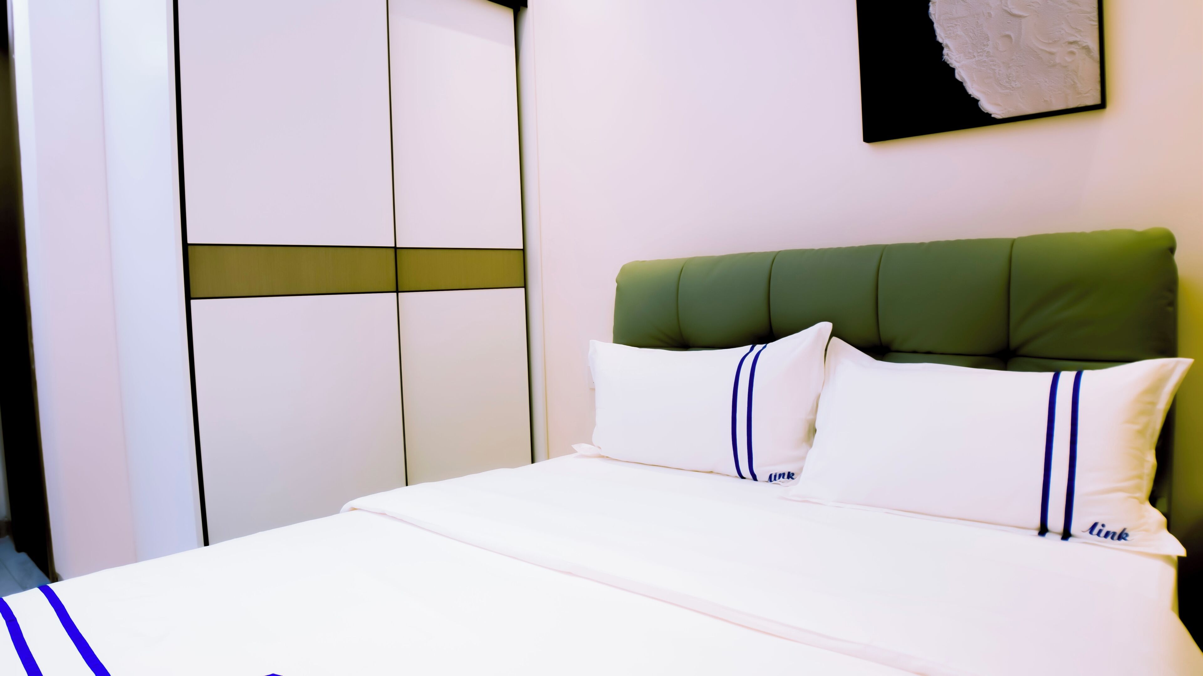 Cama com roupa de cama branca e cabeceira verde, parede branca com uma obra de arte emoldurada e porta com design verde e branco.