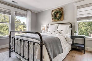 Quarto com cama, mesa de cabeceira, objeto decorativo na parede e janelas amplas.