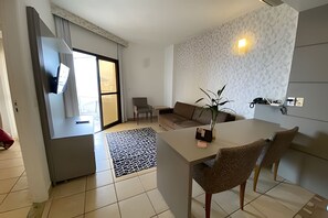 Uma sala moderna com sofá, mesa de jantar com cadeiras, pequena área de cozinha e porta de vidro deslizante para uma varanda.