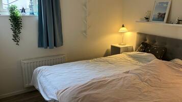 Quarto com cama, luminária, radiador, janela com cortinas e uma planta.