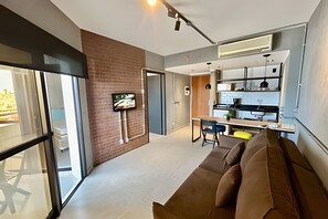 Uma sala de estar moderna com um sofá marrom, uma TV fixada na parede de tijolos e uma área de cozinha com armários e bancada.