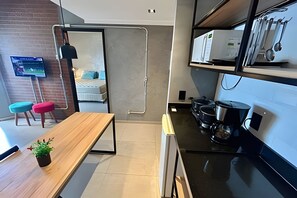 Cozinha compacta com micro-ondas, chaleira e máquina de café. Há uma pequena mesa de jantar com uma planta, e uma TV fixada na parede.