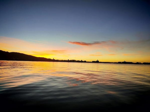 Água calma refletindo um pôr do sol, com silhuetas de edifícios no horizonte.