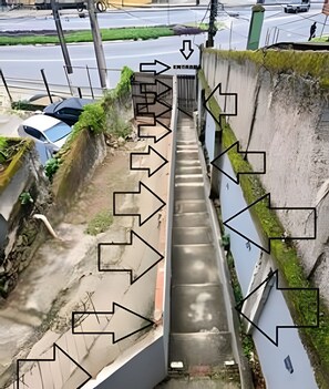 Uma trilha de concreto estreita com setas indicativas, ladeada por vegetação e uma estrada ao fundo.