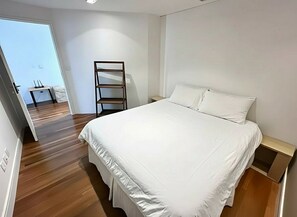 Um quarto com uma cama grande, piso de madeira, parede branca e uma porta que dá para outro cômodo.