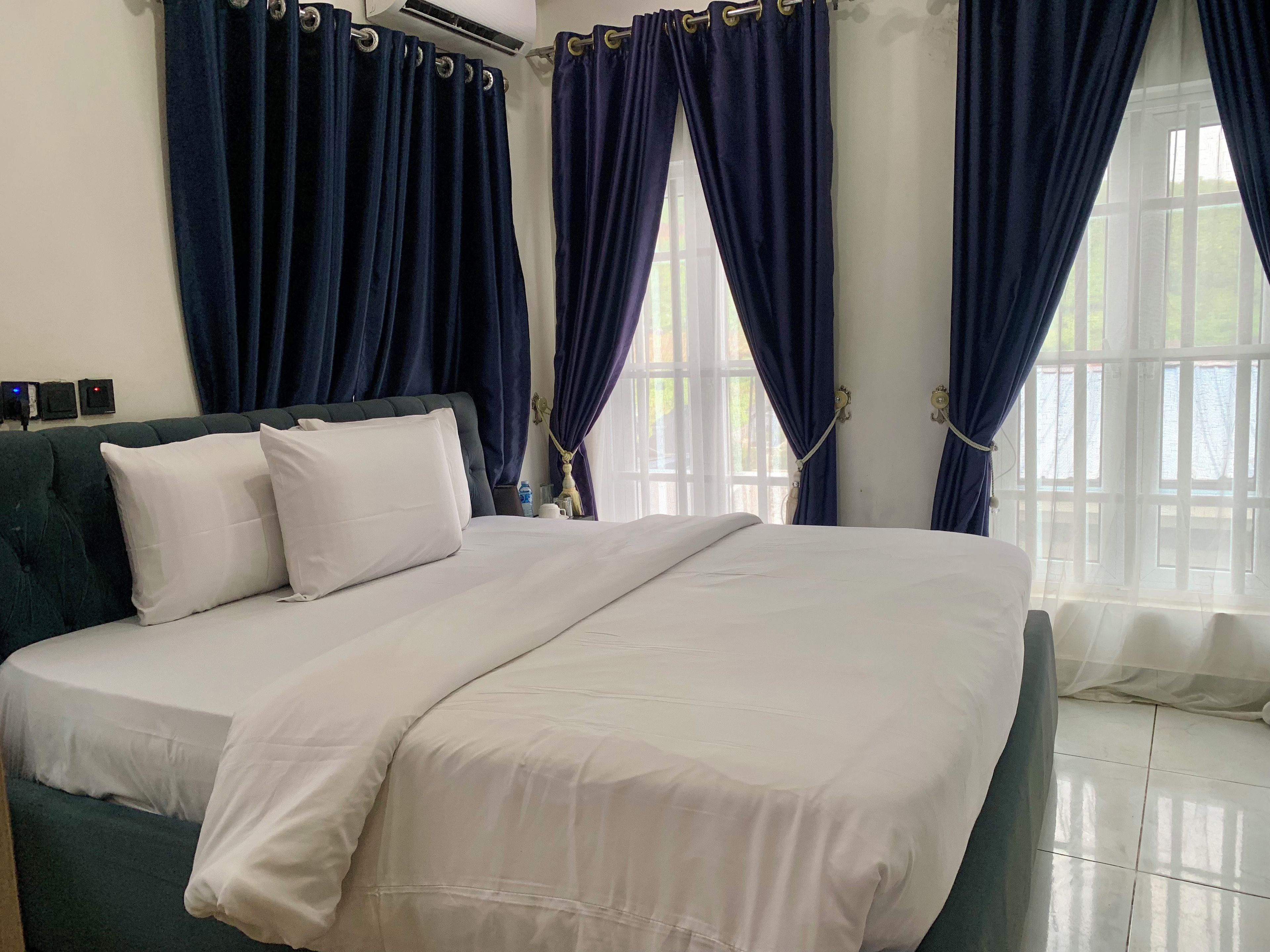Cama bem arrumada com roupa de cama e almofadas brancas, cabeceira verde escura e janela com cortinas azuis.