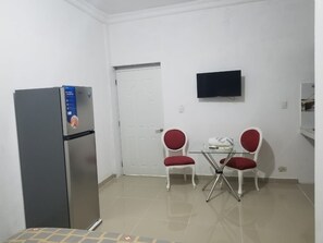 Cozinha compacta com geladeira, TV de tela plana na parede e duas cadeiras vermelhas ao redor de uma mesa pequena.
