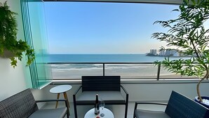 Varanda com vista para a praia e oceano. Há duas cadeiras e uma mesa pequena com uma garrafa e um copo.
