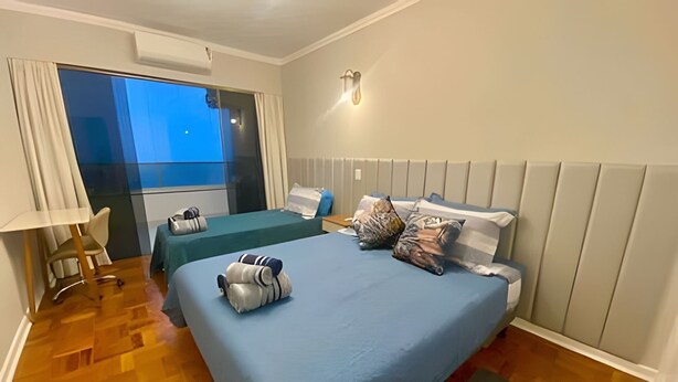 Quarto de hotel com duas camas, uma escrivaninha, uma cadeira, janela com cortinas e luminária na parede.
