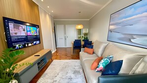 Uma sala de estar moderna com uma TV de tela plana, um sofá, uma mesa de centro e uma área de jantar ao fundo.