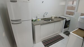 Cozinha com armários brancos, bancada de granito, pia, geladeira e janela com persianas.