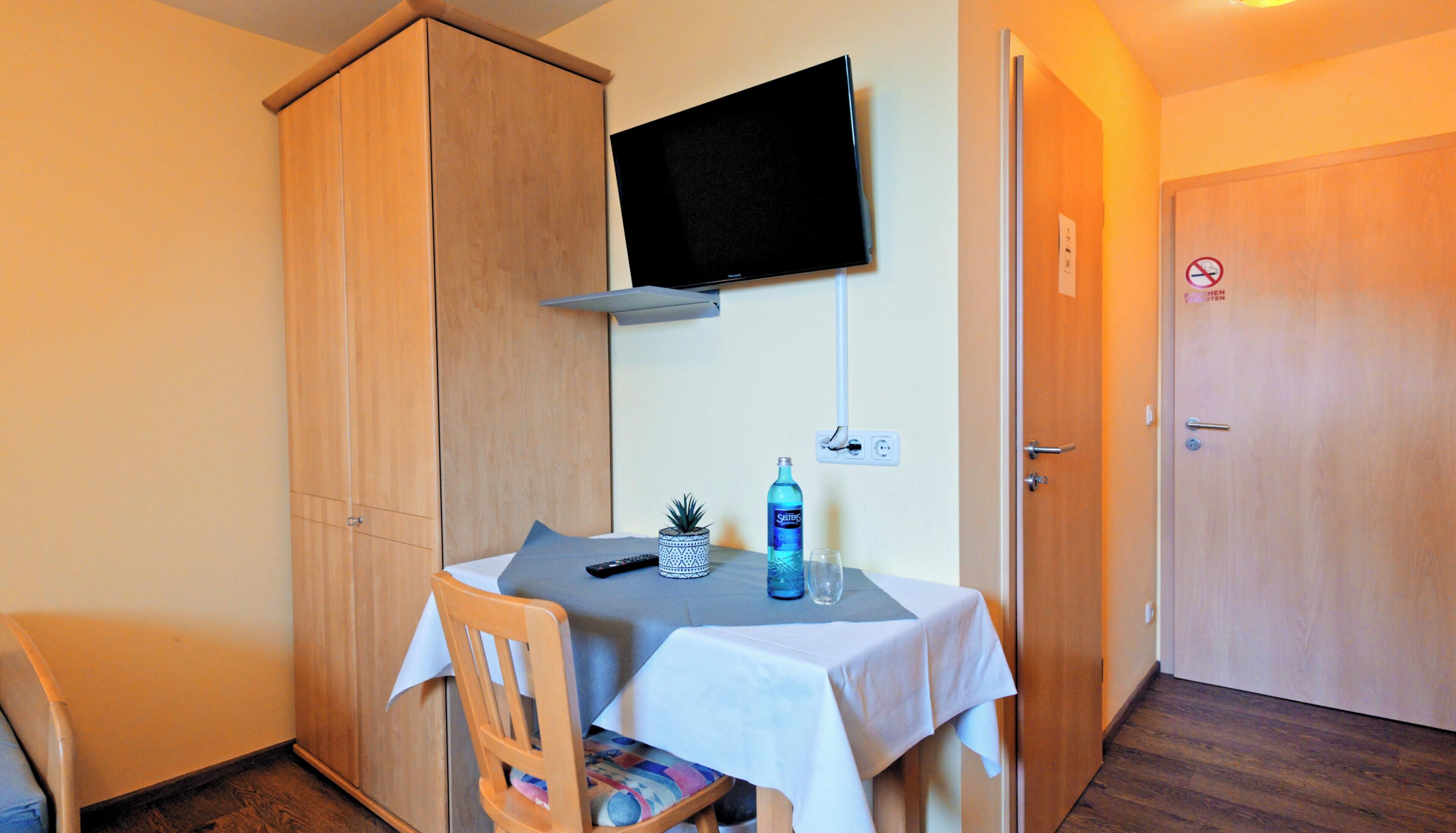 Um quarto pequeno com uma mesa, uma cadeira, uma televisão na parede e uma porta.