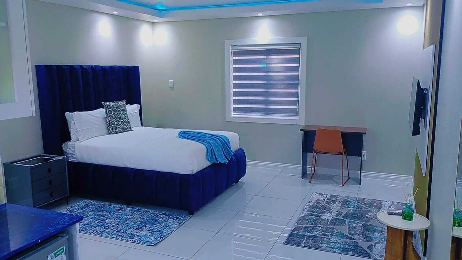 Quarto com cama, mesa de cabeceira, escrivaninha, cadeira, janela e uma pequena mesa com uma garrafa.