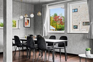 Uma área de jantar moderna com uma mesa grande, seis cadeiras, luminárias pendentes e vista para um edifício de tijolos.