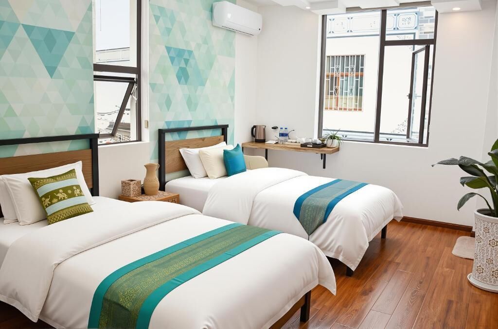 Quarto de hotel com duas camas, uma janela, uma prateleira com objetos e um vaso de planta.