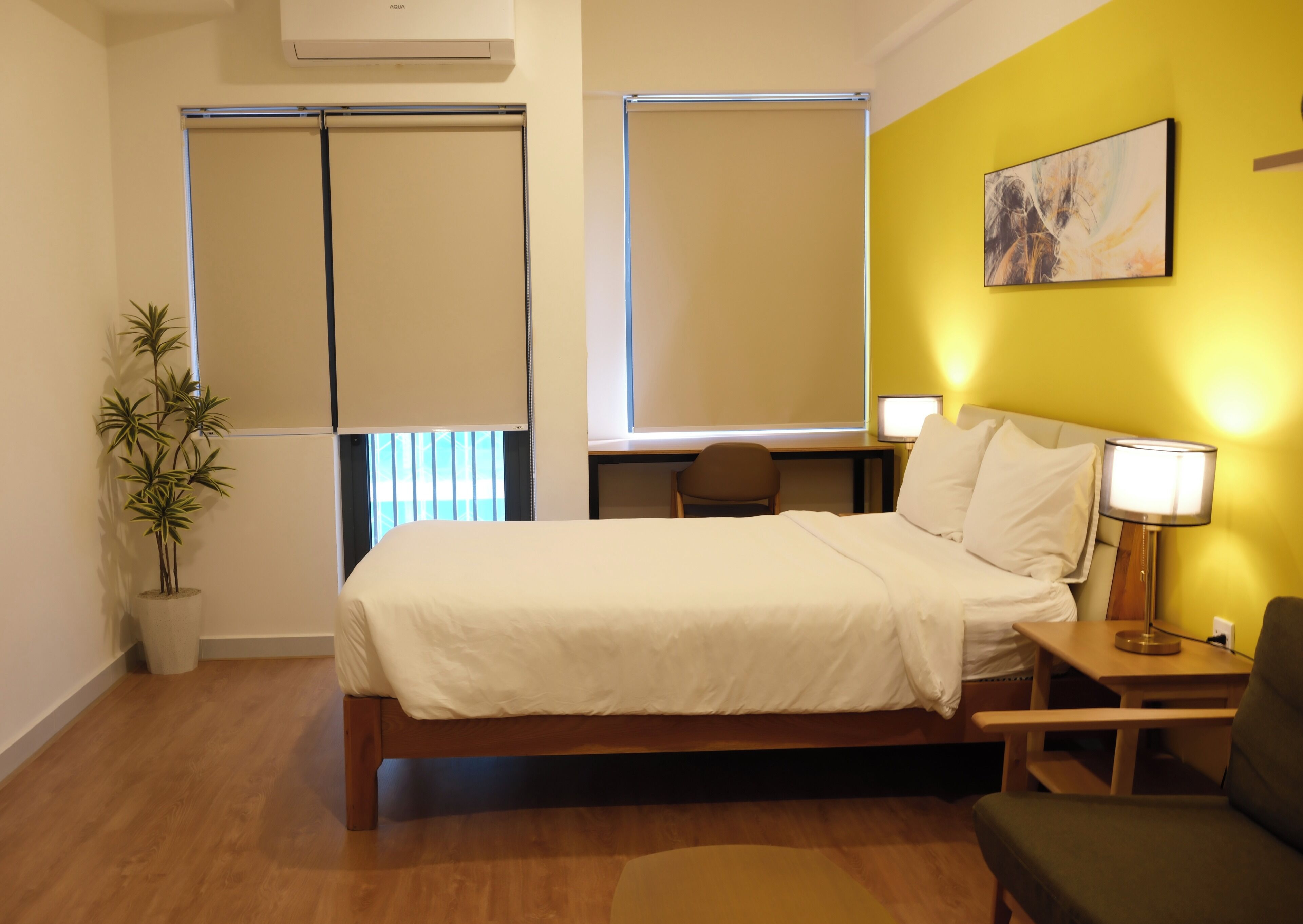 Quarto de hotel com cama, mesa de cabeceira, abajur, cadeira, escrivaninha e planta em vaso.