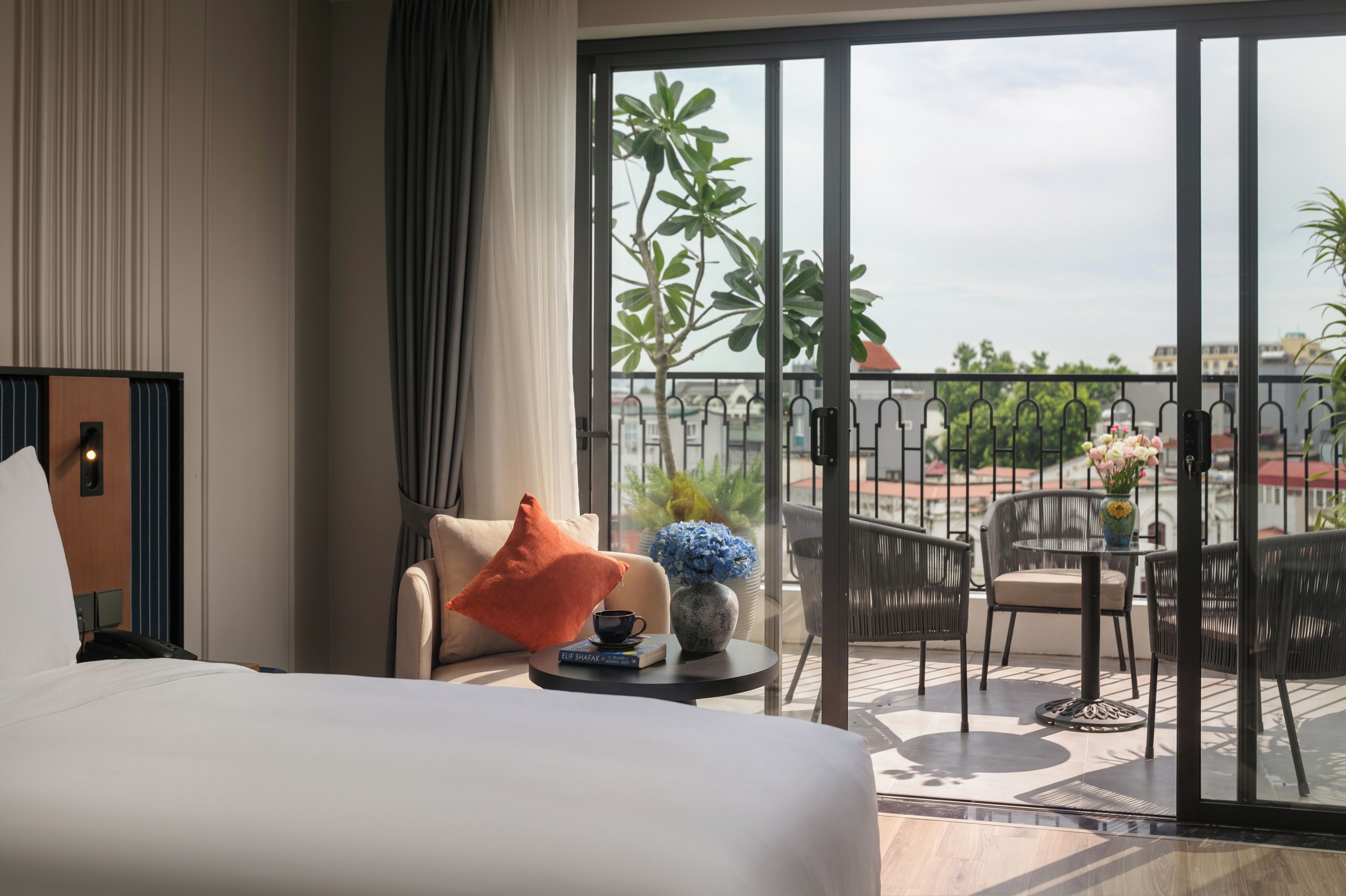 Quarto de hotel com cama, sofá, mesa com um vaso de flores e varanda com vista para a cidade.