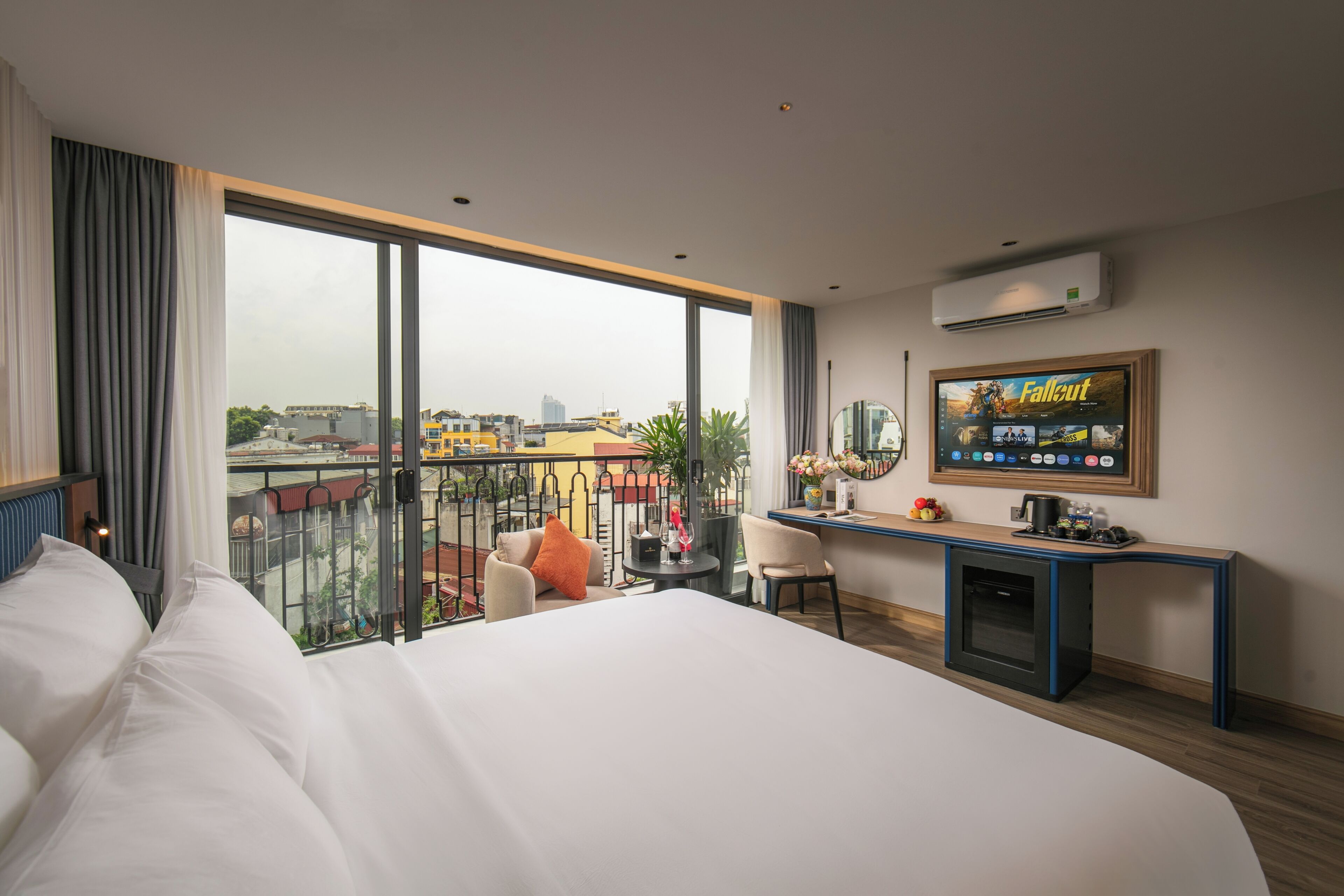 Quarto de hotel com uma cama grande, uma escrivaninha com um computador, uma TV exibindo um jogo e uma varanda com vista para a cidade.
