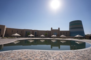 Piscina com deck ladrilhado, guarda-sóis e um edifício com uma torre imponente ao fundo.