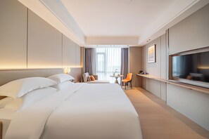 Quarto de hotel com uma cama grande, televisão, mesa pequena com uma cadeira e vista para a cidade pela janela.