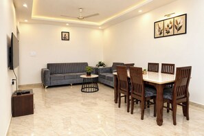 Uma sala de estar moderna com um sofá, uma mesa de jantar com cadeiras, uma pequena mesa redonda com uma planta, um ventilador de teto, iluminação embutida e um quadro na parede.