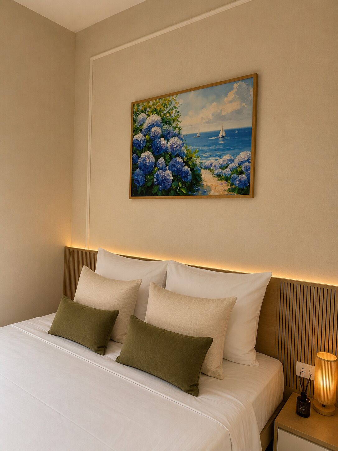 Quarto com cama, almofadas, mesa de cabeceira com abajur e pintura de paisagem costeira.
