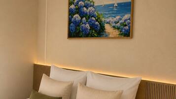 Quarto com cama, almofadas, mesa de cabeceira com abajur e pintura de paisagem costeira.